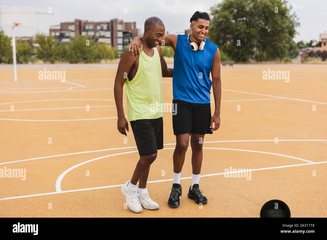 Smiling father and son standing with arms around at basketball court ...