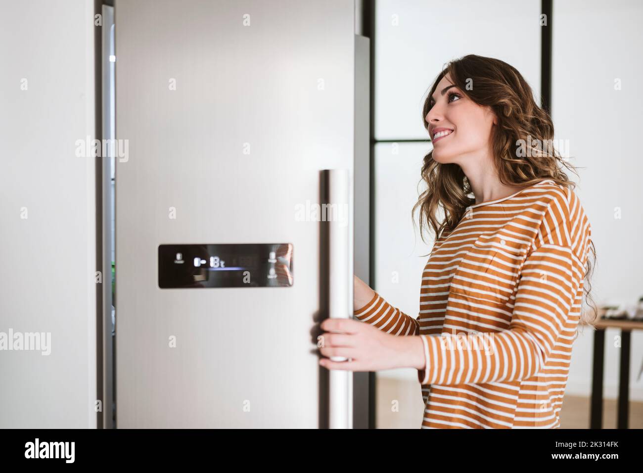 Smiling young woman in kitchen opening fridge Stock Photo - Alamy