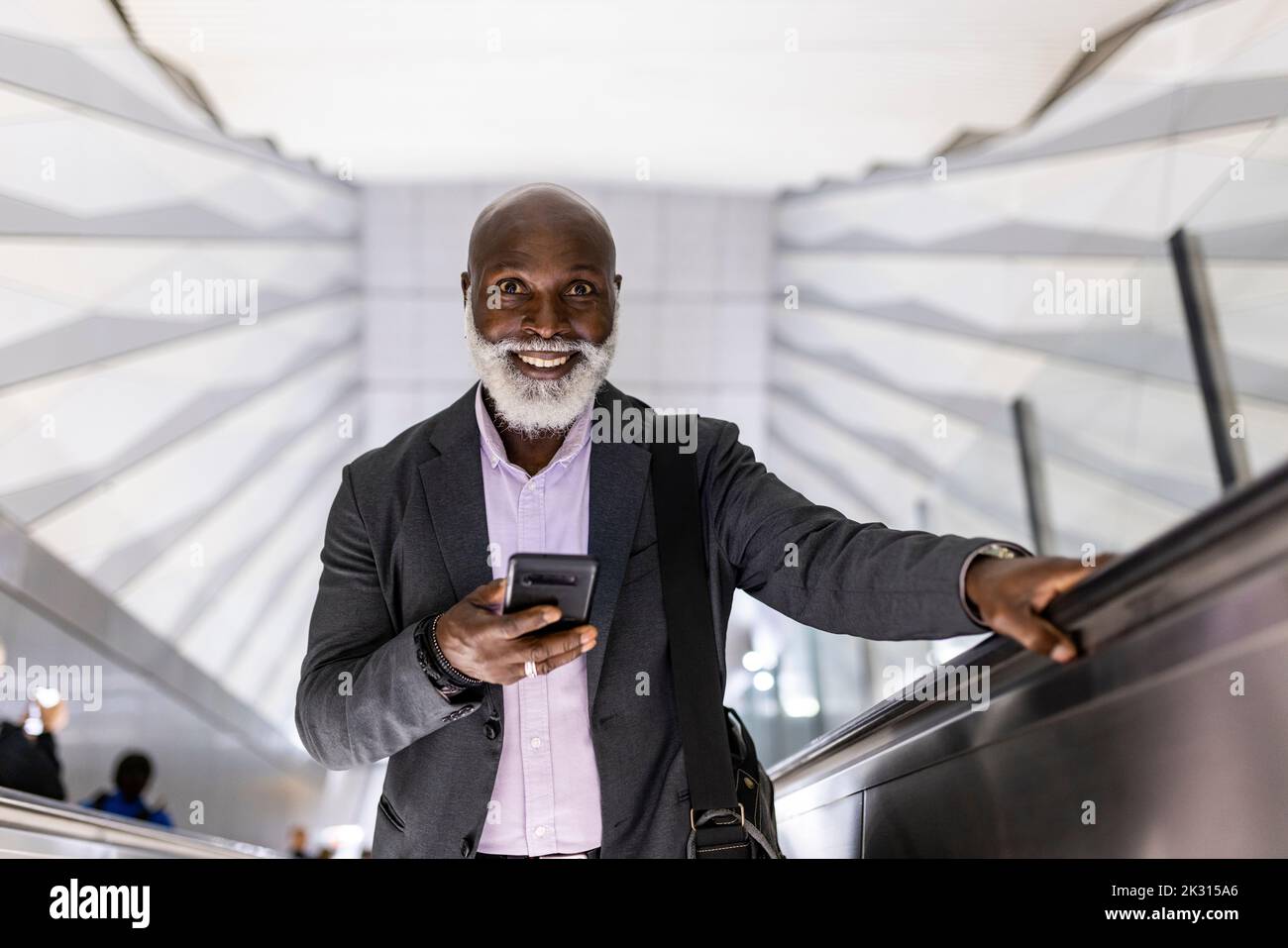Smiling bald commuter with smart phone on escalator at subway station ...