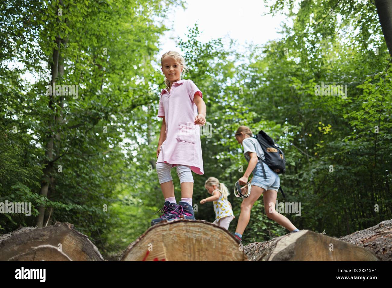 Smiling girl standing on tree trunk with mother and daughter walking in ...