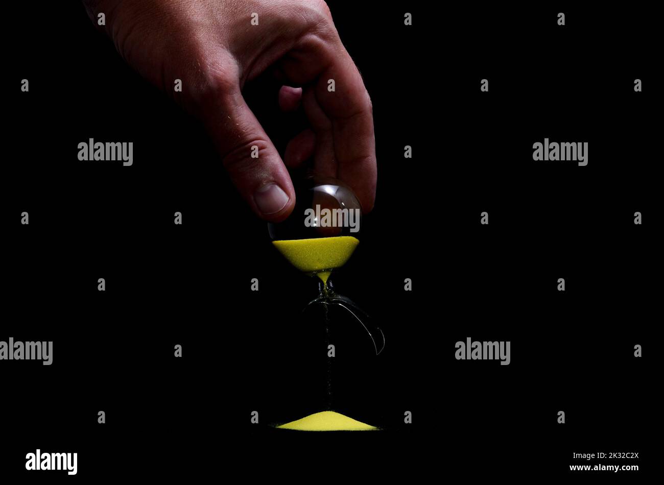 A hand holding a broken hourglass with yellow sand on black background ...