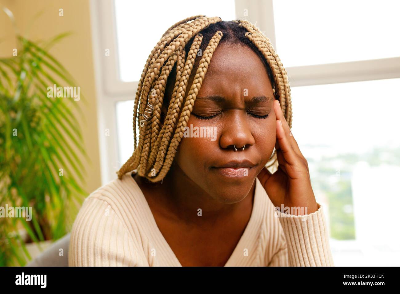 worried woman with closed eyes sitting near window and green plant at ...