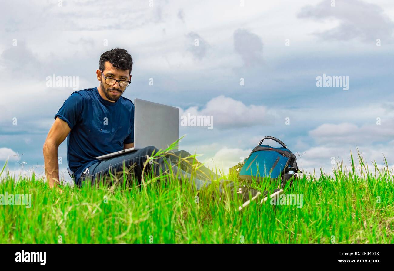Young man sitting with laptop and backpack in the field. Man sitting in ...