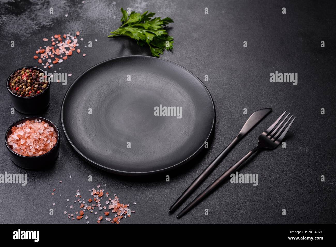 An empty black plate on a dark concrete background with spices, herbs ...