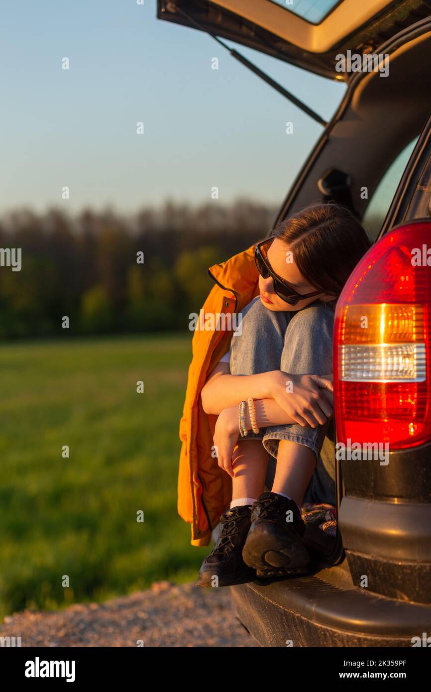 Pretty sad teenage girl with sun glasses sitting alone in a car trunk ...