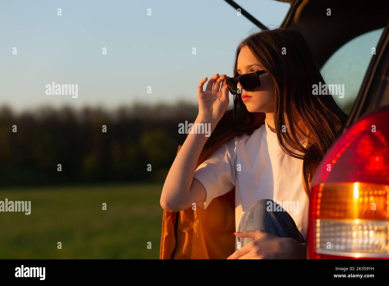 Pretty sad teenage girl with sun glasses sitting alone in a car trunk ...