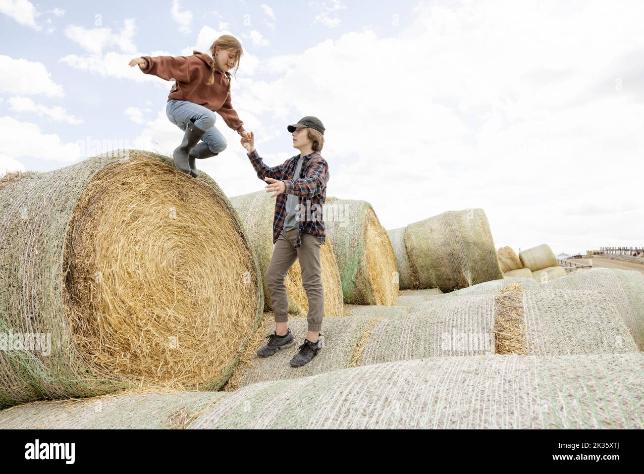 Brother helping sister jumping from rolled hay bale on farm Stock Photo ...