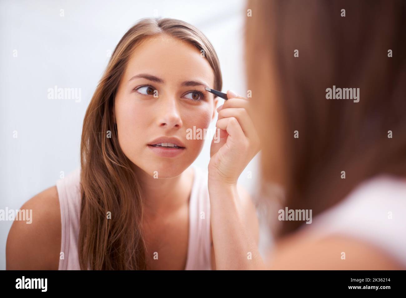 Creating the perfect arch. A young woman tweezing her eyebrows in front ...