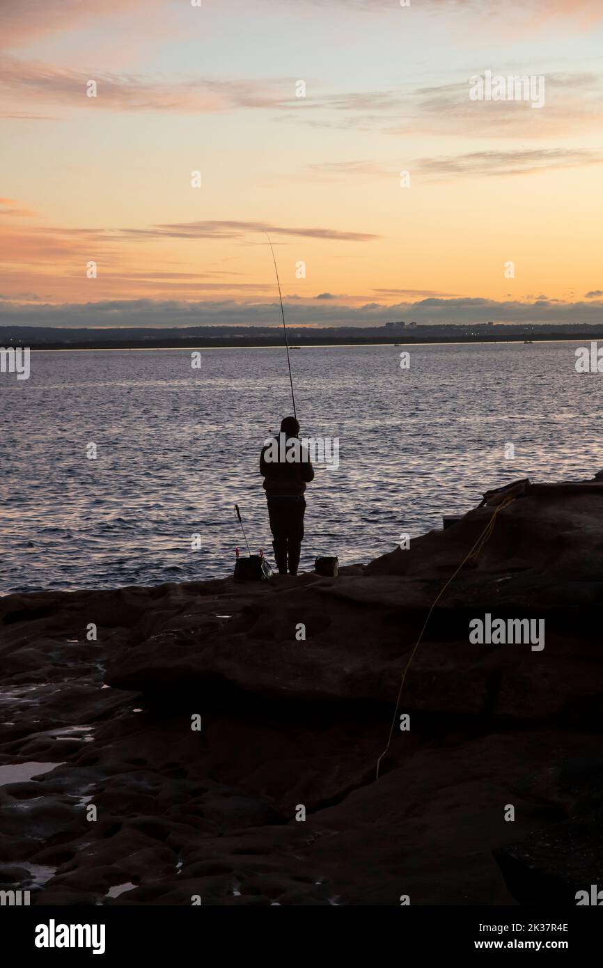 A silhouette shot of a person standing on a rocky beach while fishing ...