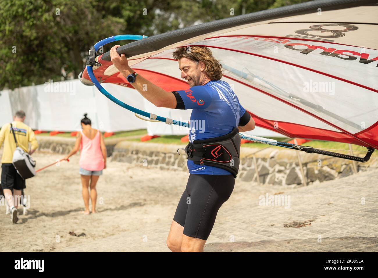 A man carries a white windsurfing sail across a beach to the sea at the ...