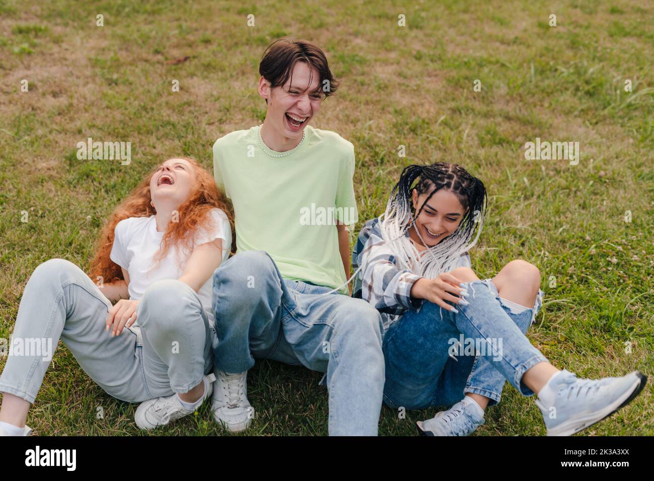 Happy young three best friends having fun together outdoors sitting on grass in public city park ...