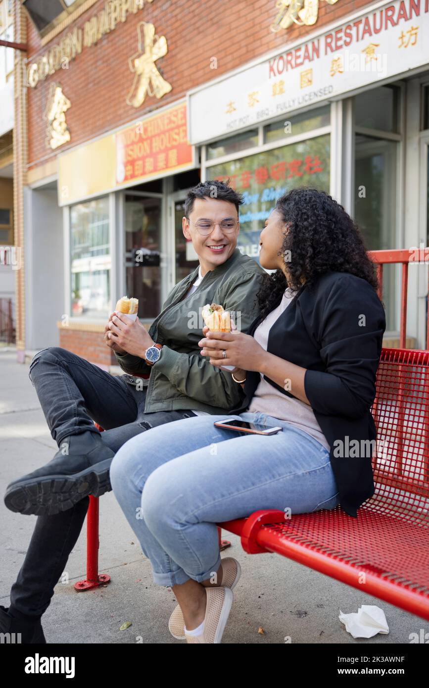 Young couple eating sandwiches on urban bench Stock Photo - Alamy