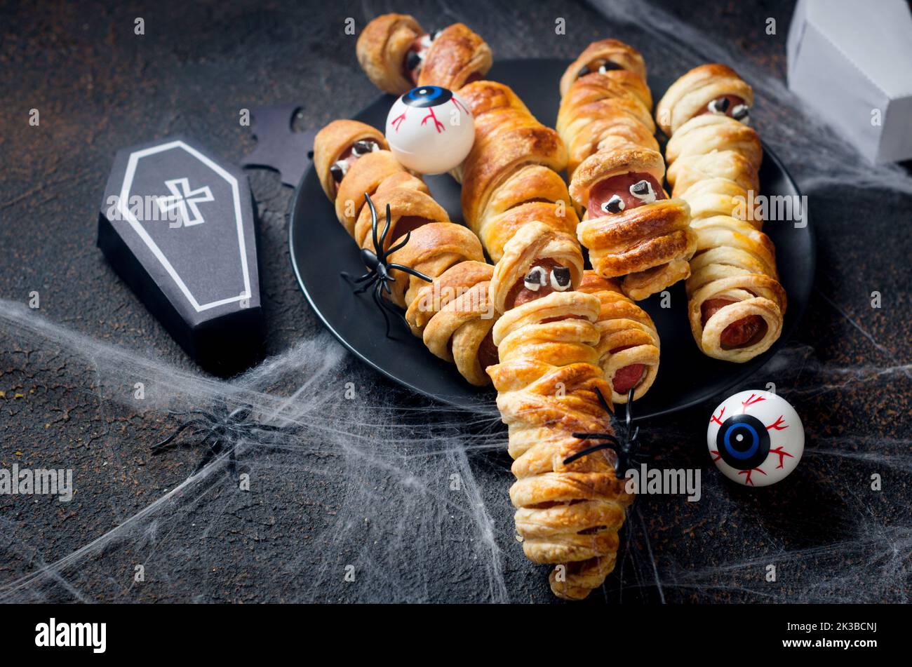 Spooky sausage mummies in dough in black plate with graves on dark ...