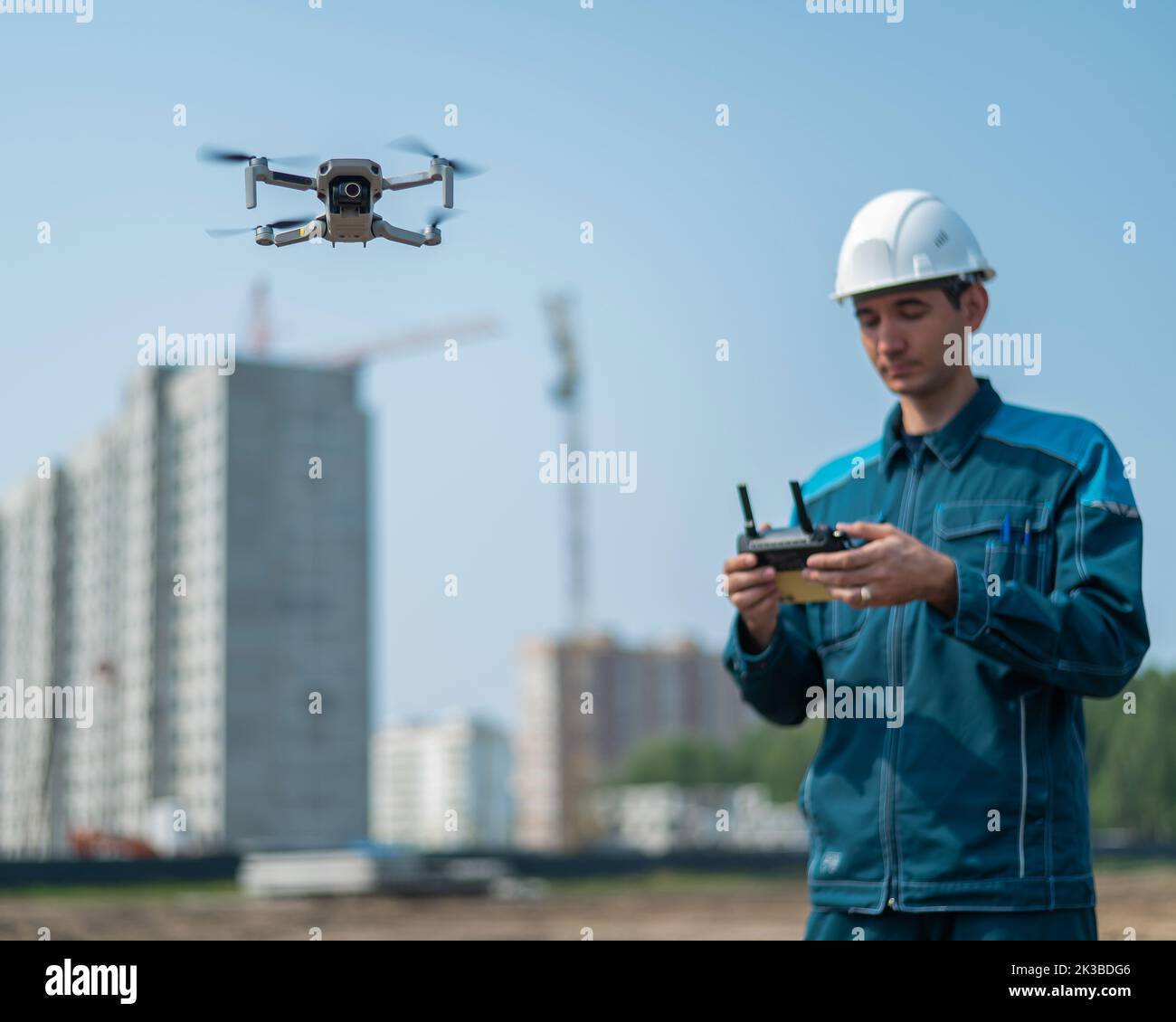 A man in a helmet and overalls controls a drone at a construction site ...