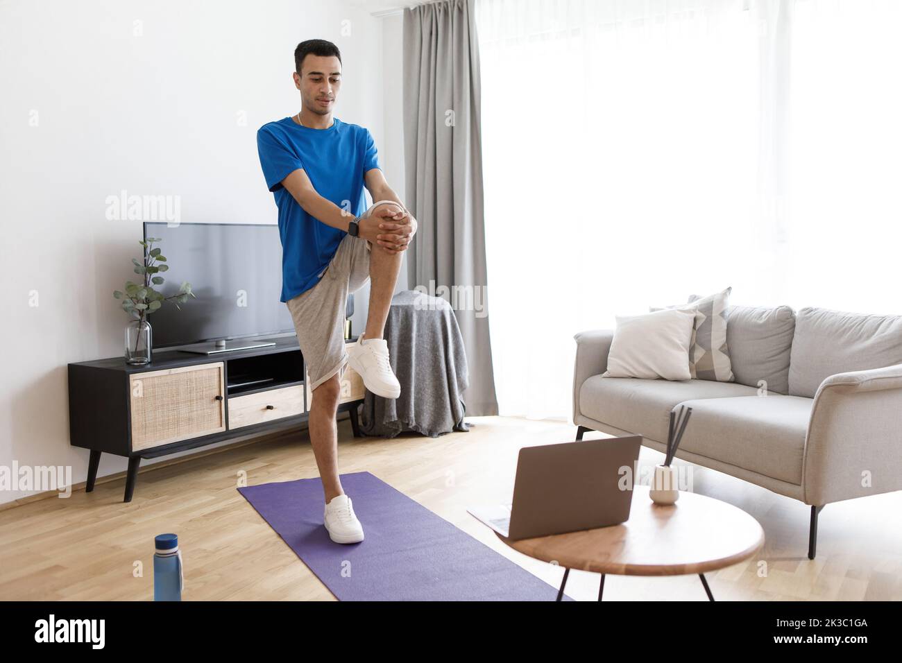Home workout routine. Young arab man stretching and doing standing knee ...