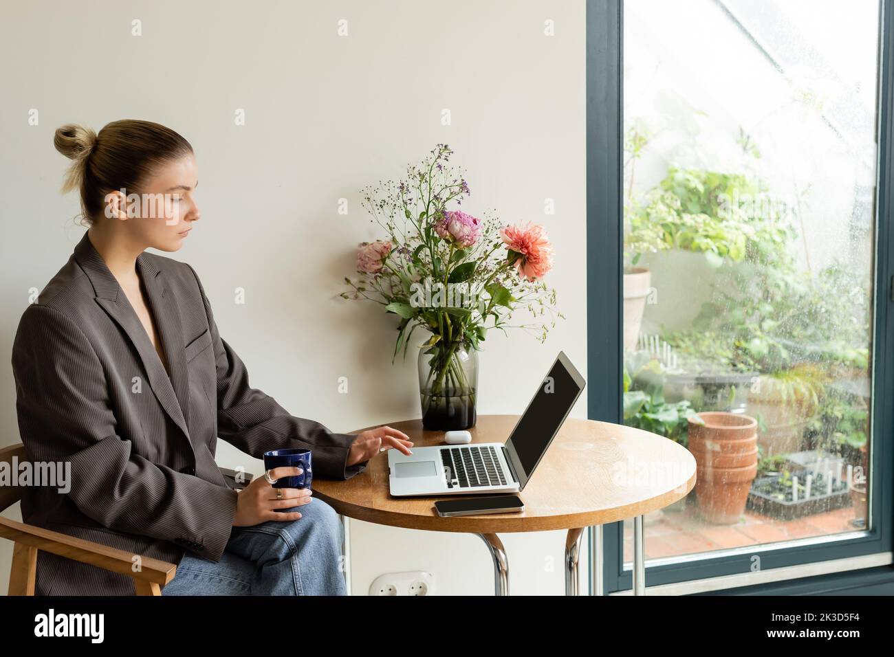 Side view of woman in jacket holding cup and using device near flowers ...