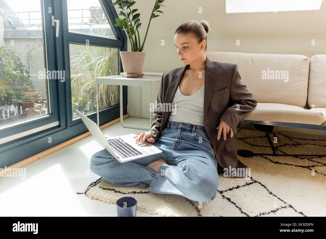 Young freelancer in jacket using laptop near cup on carpet at home ...