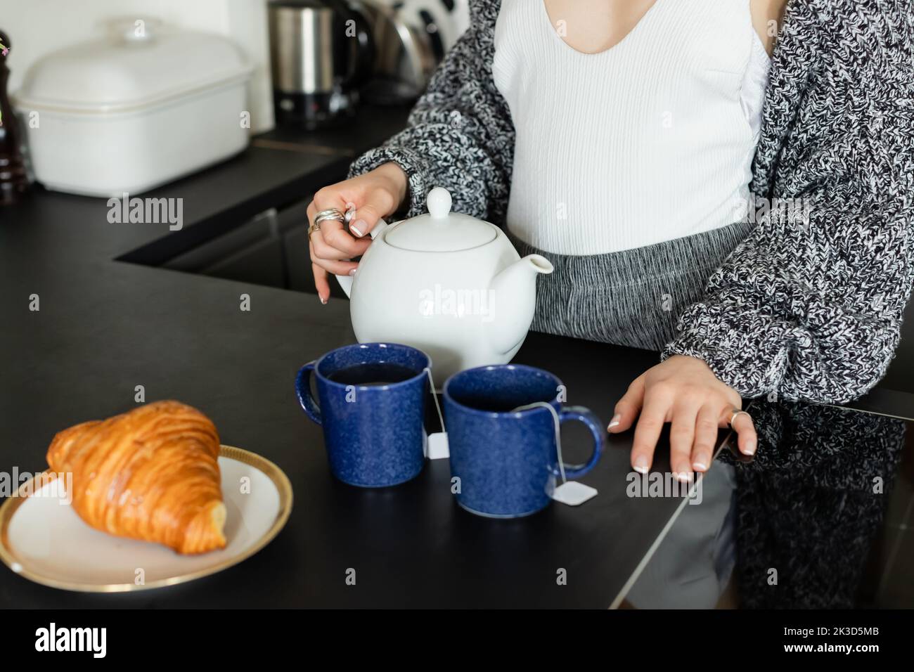 Cropped view of woman in knitted cardigan holding teapot near cups and ...