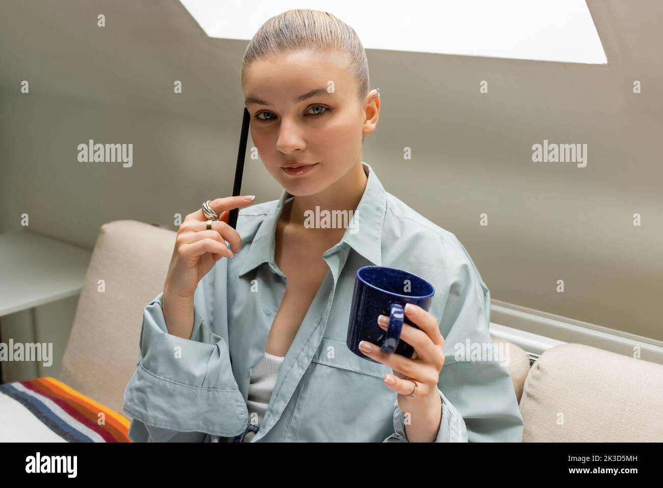 Young woman holding pen and cup while looking at camera on couch in ...