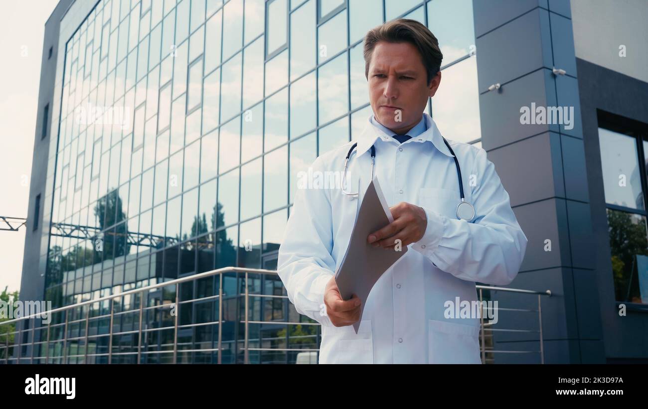 serious doctor in white coat holding folder with documents near ...