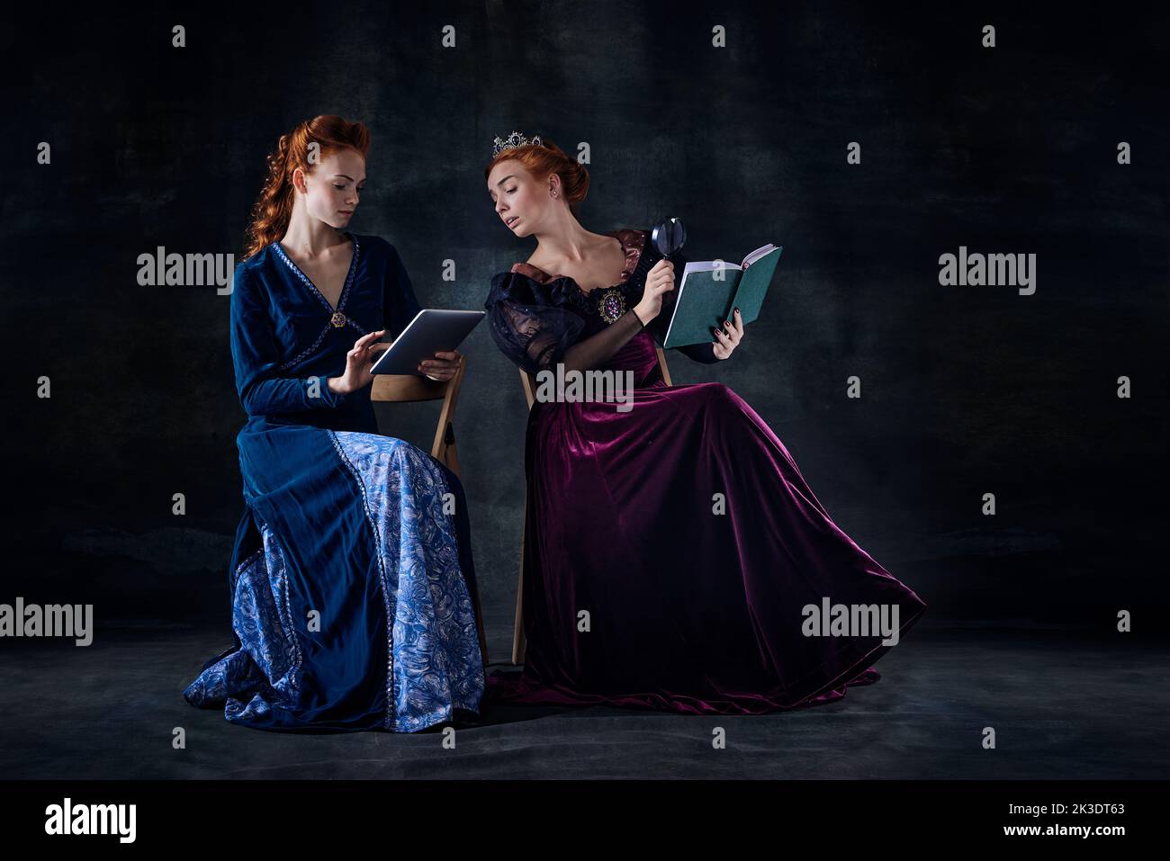 Portrait of two beautiful women in image of queens reading isolated over dark background ...