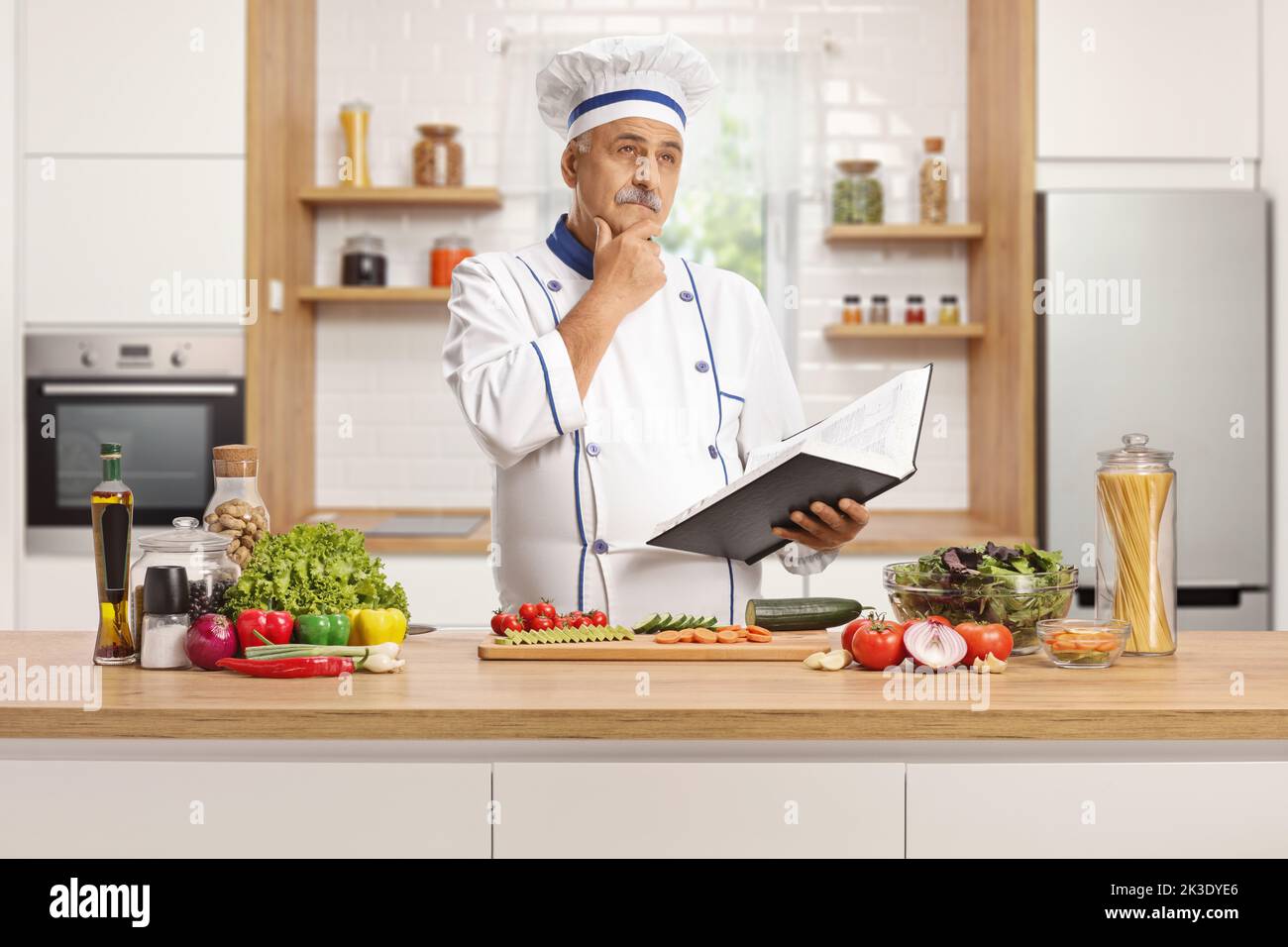 Mature male chef holding a cook book and thinking inside a kitchen ...