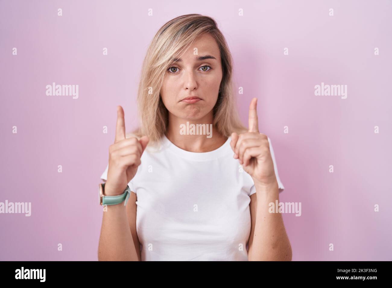 Young blonde woman standing over pink background pointing up looking ...
