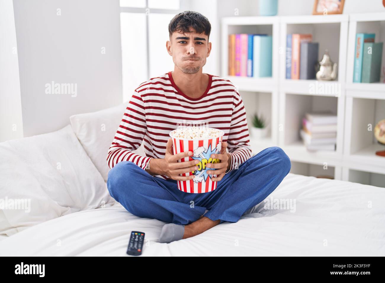 Young hispanic man eating popcorn sitting on the bed watching a movie ...
