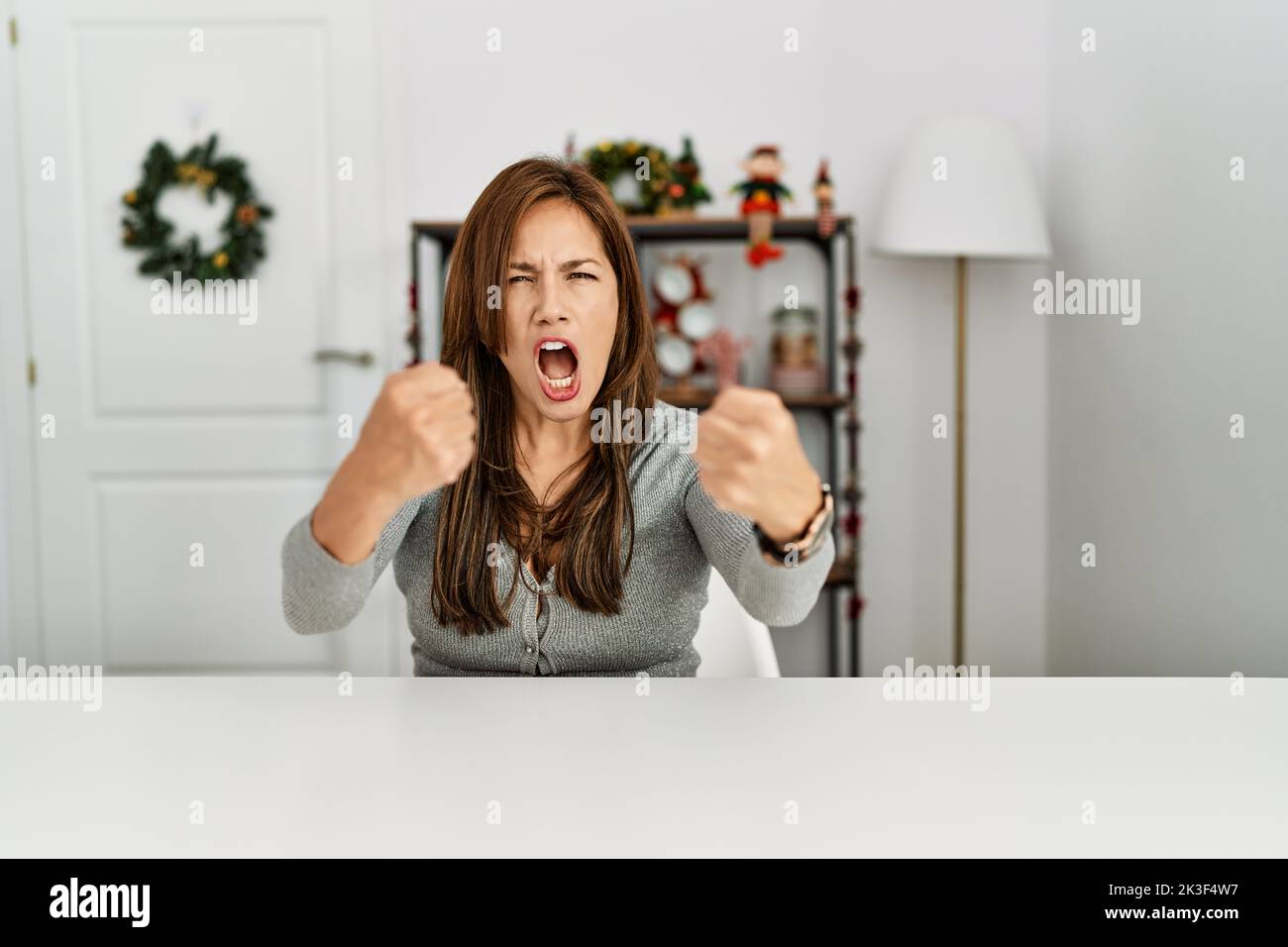 Young latin woman sitting on the table by christmas decor angry and mad ...