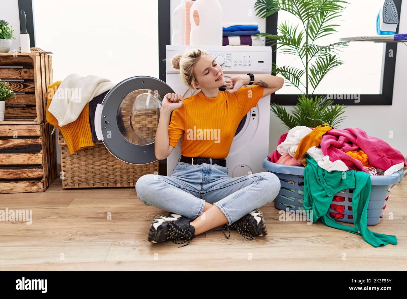 Young blonde woman doing laundry sitting by washing machine stretching ...