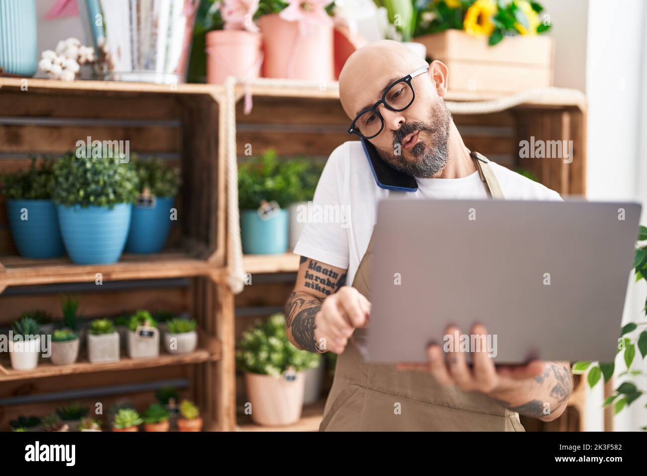 Young bald man florist talking on smartphone using laptop at florist ...