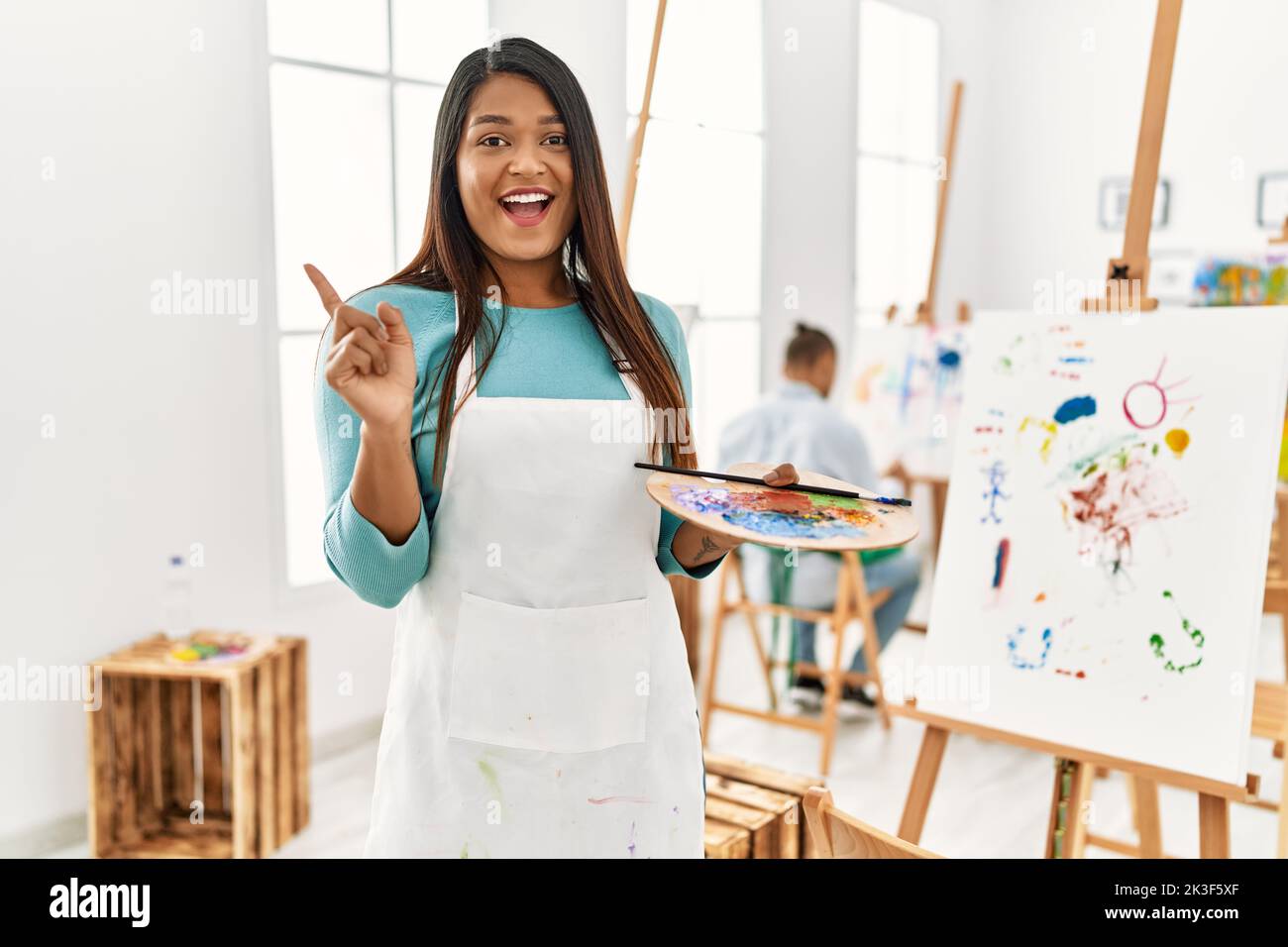 Young latin woman standing at art studio smiling happy pointing with ...