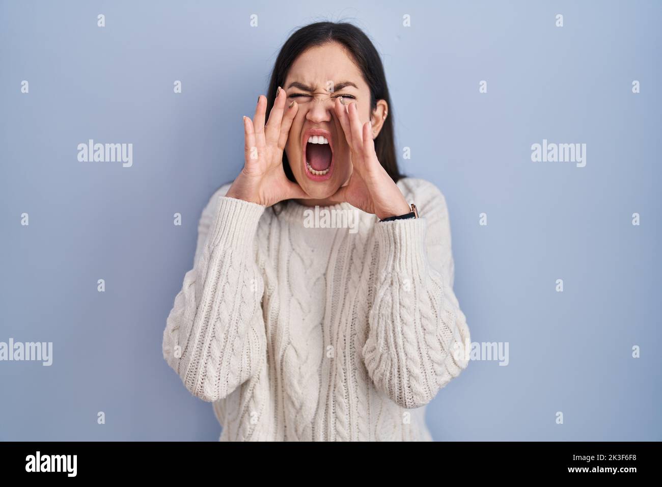 Young brunette woman standing over blue background shouting angry out ...