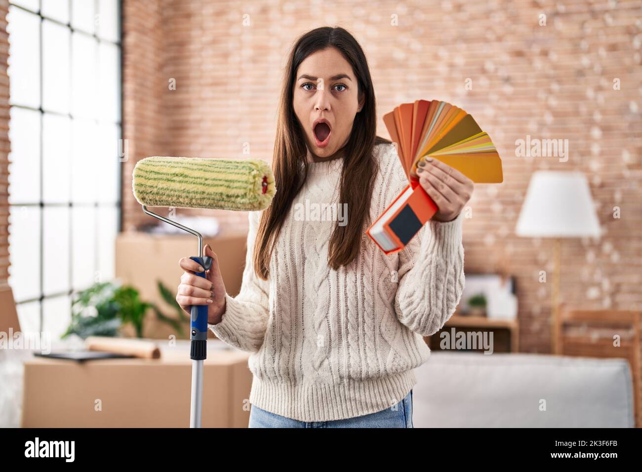 Young brunette woman holding roller painter and paint samples in shock ...