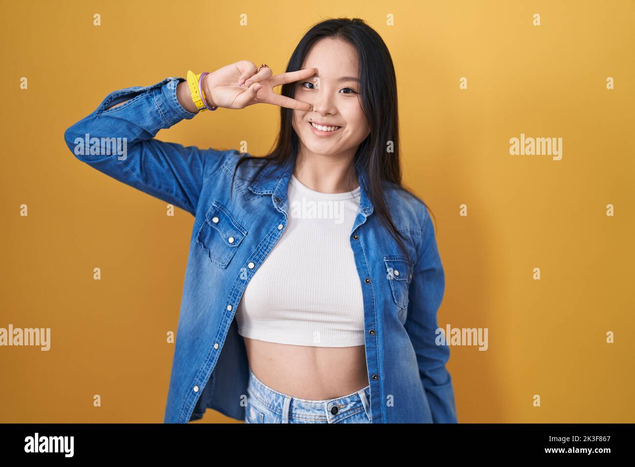 Young asian woman standing over yellow background doing peace symbol ...