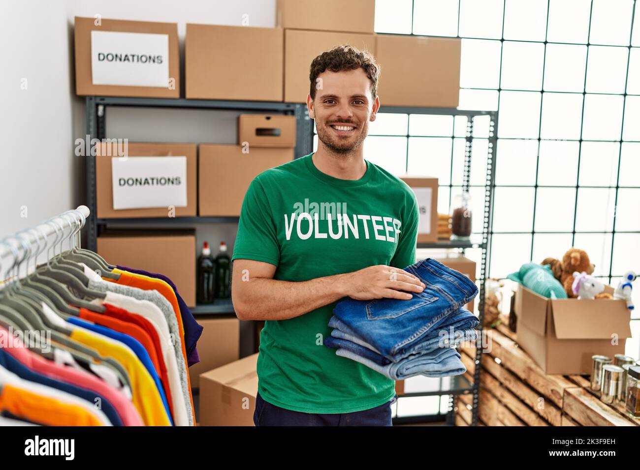 Young hispanic man wearing volunteer uniform holding folded jeans at ...