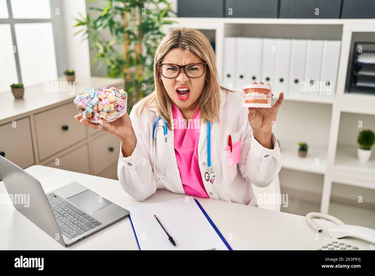 Young hispanic dentist woman holding denture and sweets angry and mad ...