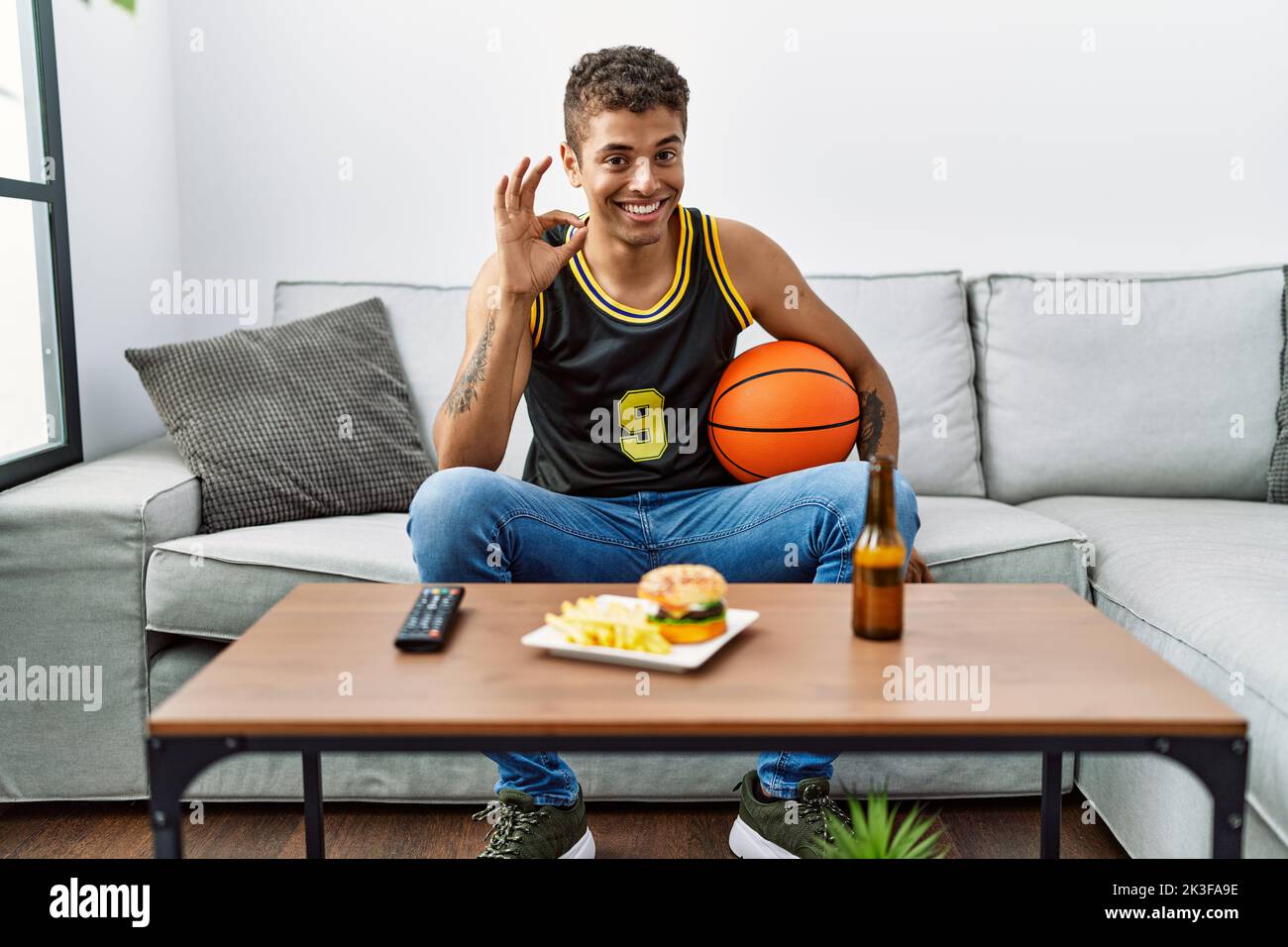 Young handsome hispanic man holding basketball ball cheering tv game ...