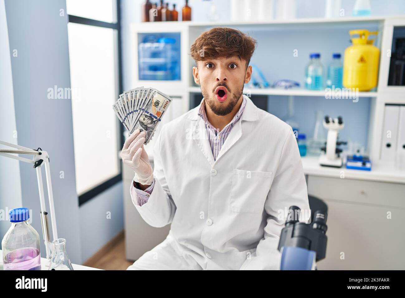 Arab man with beard working at scientist laboratory holding money scared and amazed with open ...