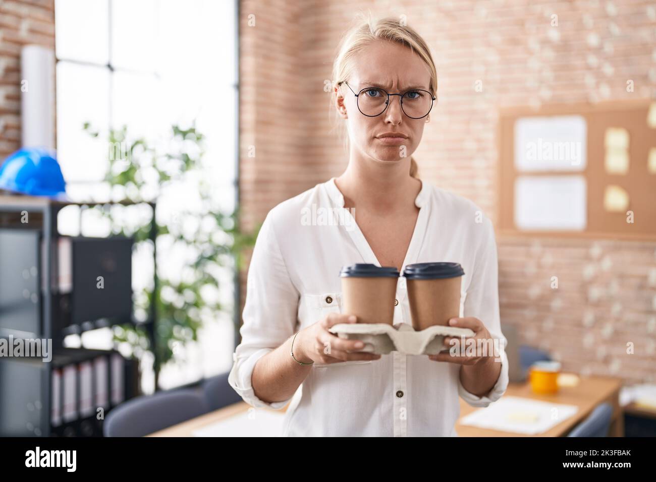 Young caucasian woman working at the office holding coffee cups skeptic ...