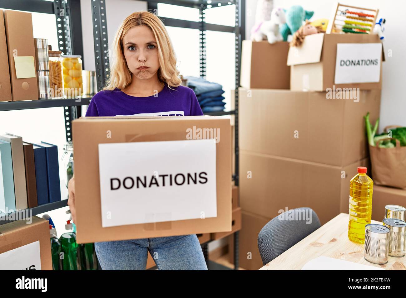 Beautiful blonde woman volunteer holding donations box puffing cheeks ...