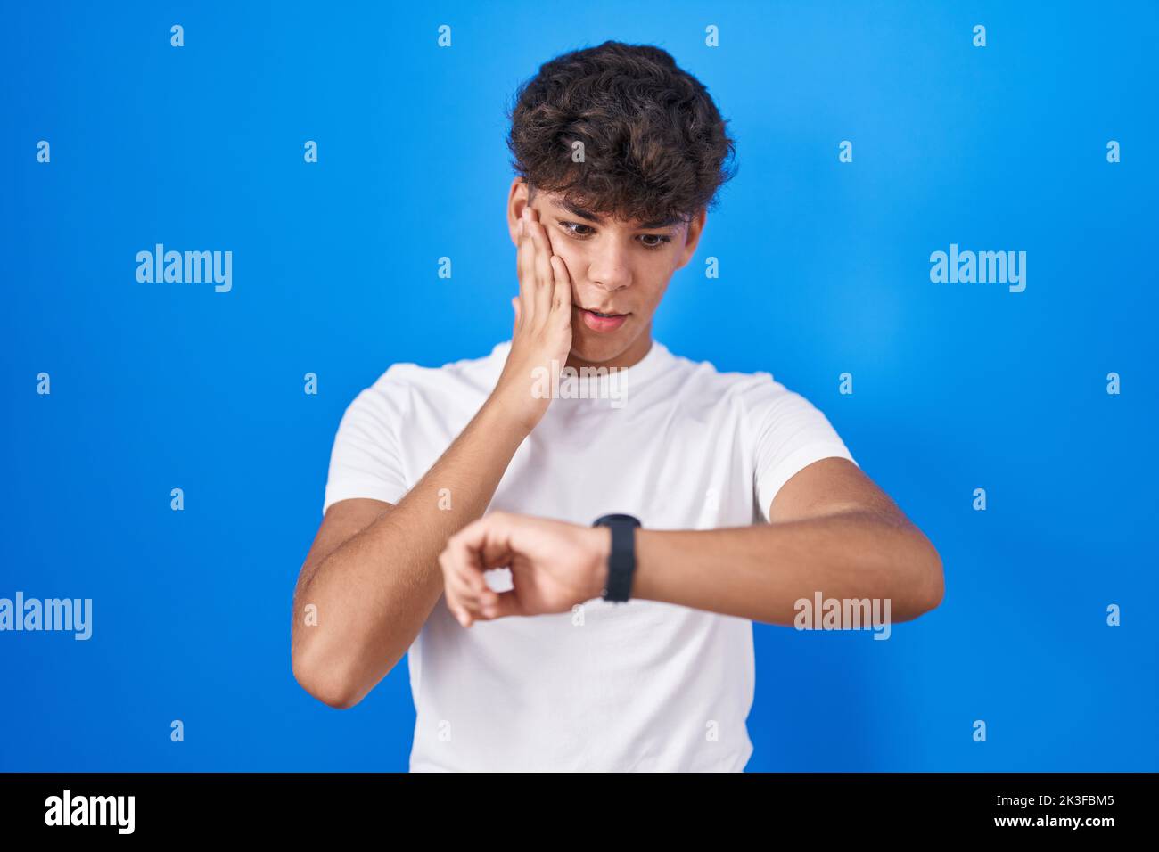Hispanic teenager standing over blue background looking at the watch ...