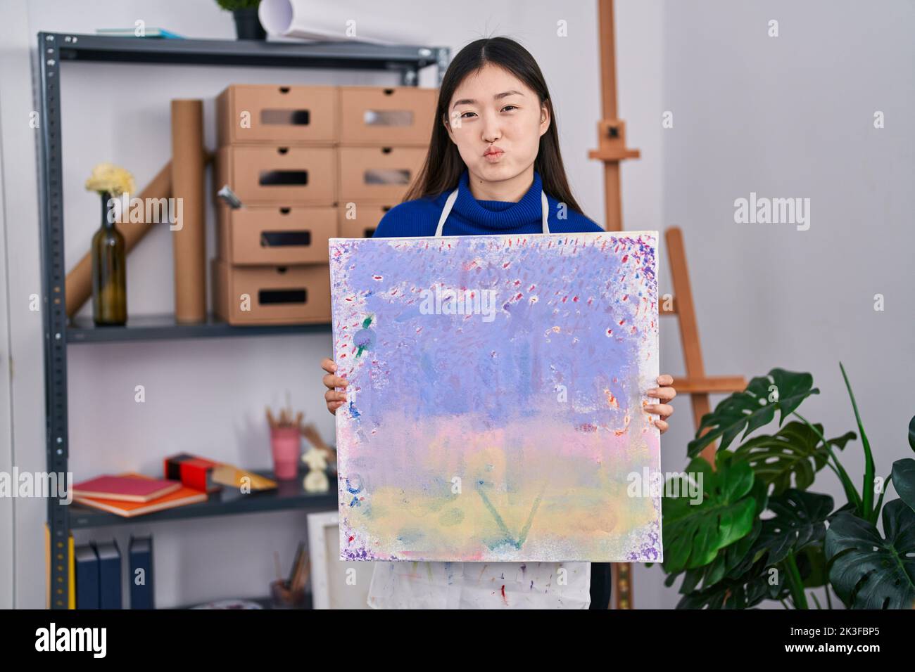 Chinese young woman holding canvas at art studio puffing cheeks with ...