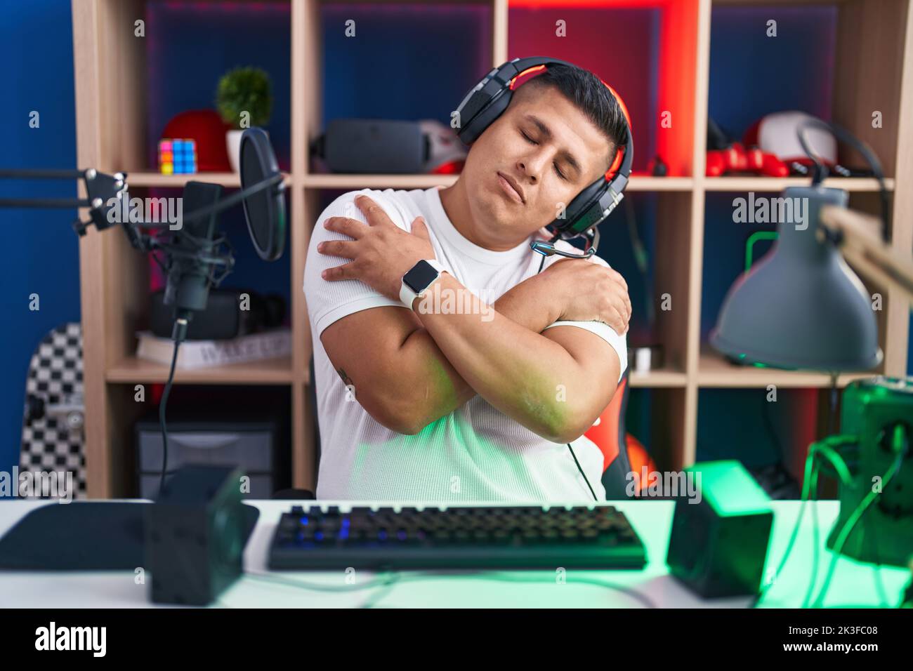 Young hispanic man playing video games hugging oneself happy and ...
