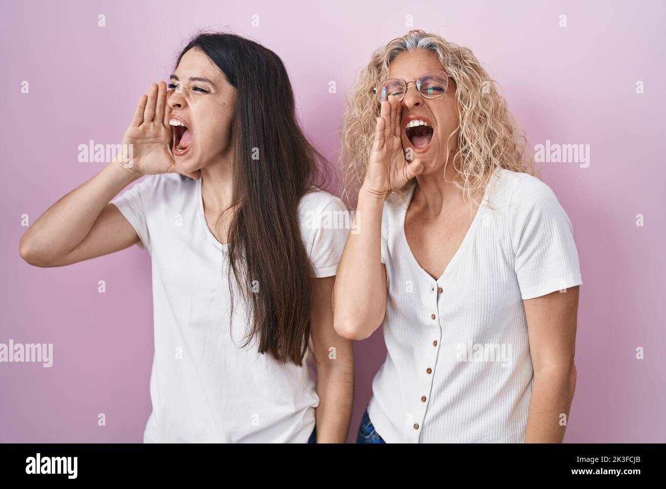 Mother and daughter standing together over pink background shouting and ...