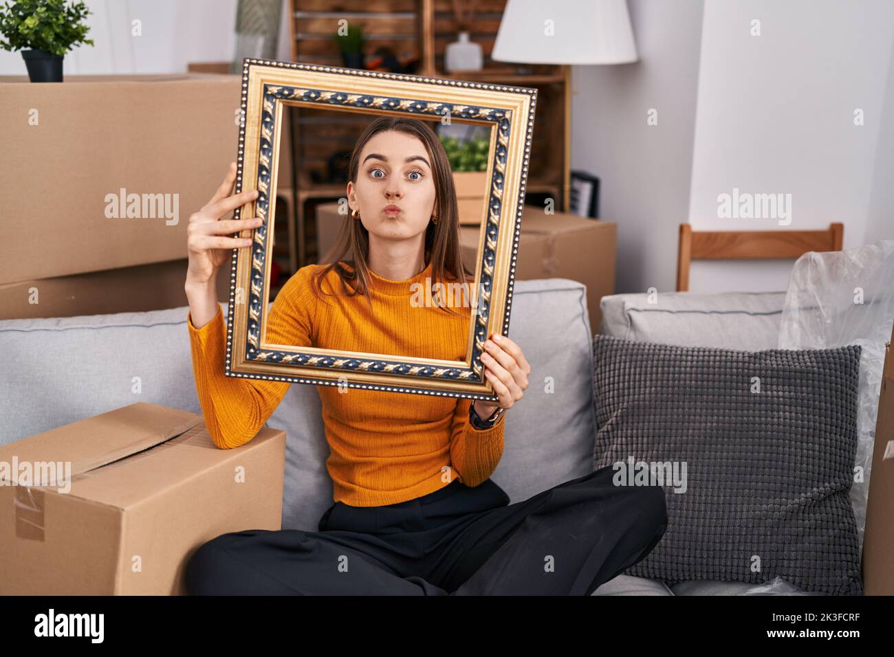 Young caucasian woman at new home holding empty frame puffing cheeks ...