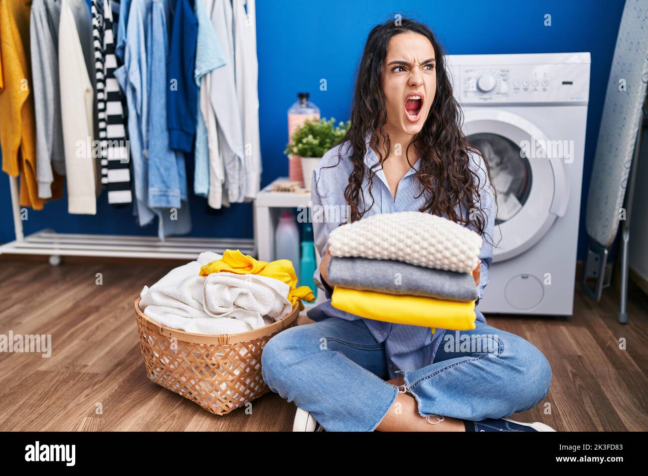 Young brunette woman holding clean laundry angry and mad screaming frustrated and furious ...