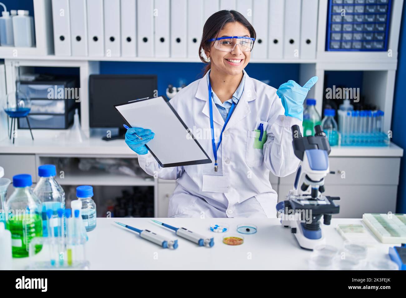 Hispanic young woman working at scientist laboratory pointing to the back behind with hand and ...