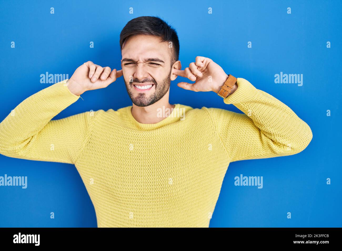 Hispanic man standing over blue background covering ears with fingers ...