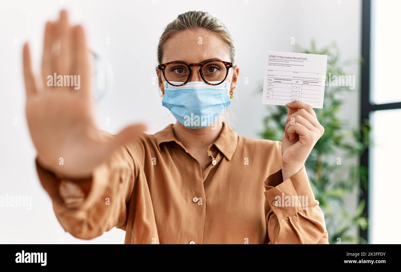 Young blonde woman holding covid record card with open hand doing stop ...